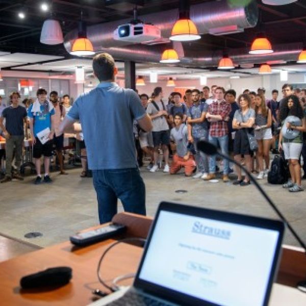 A color photo of a person speaking with the a large group of people gathered around him.